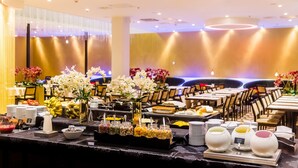 Disposição de buffet com diversos alimentos, incluindo frutas, doces e acompanhamentos, dispostos em mesas com cadeiras.