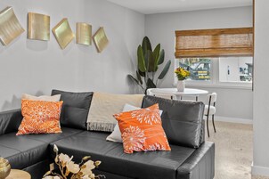 Uma sala de estar moderna com um sofá de couro preto, almofadas estampadas em laranja e um vaso branco com flores.