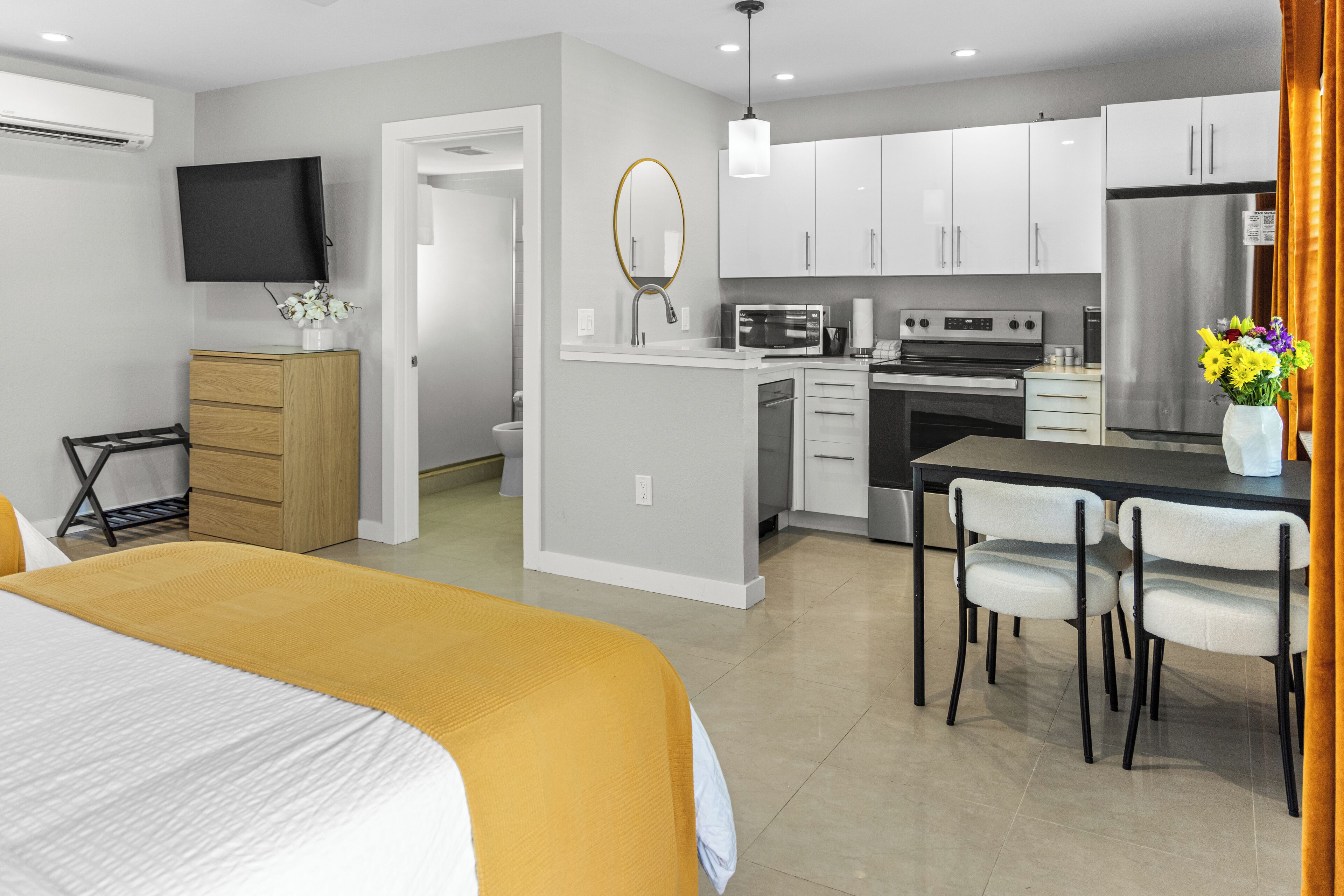 Quarto de hotel moderno com uma cama grande, uma kitchenette pequena, uma área de refeições com duas cadeiras e uma TV fixada na parede.