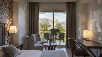 Quarto de hotel com cama, duas poltronas, escrivaninha e varanda com vista para as montanhas.