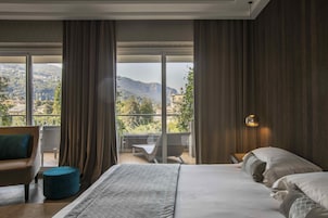 Quarto com uma cama grande, uma cadeira, um banquinho pequeno e vista para uma varanda com cadeira e mesa.