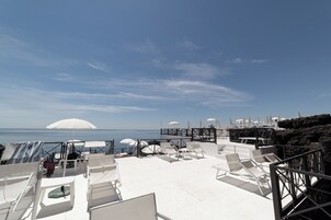 Um terraço à beira-mar com cadeiras de lounge brancas e guarda-sóis, com vista para o oceano.