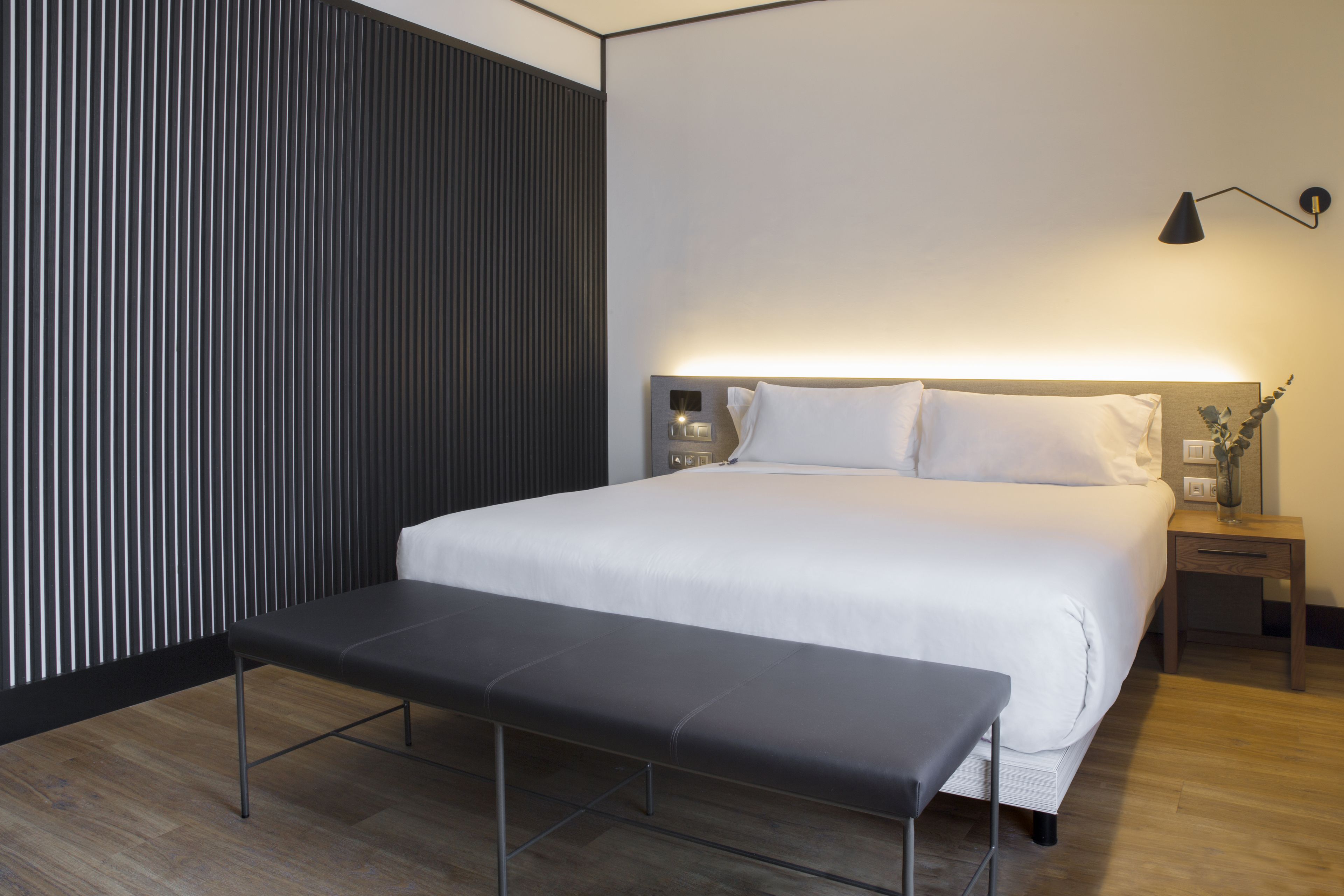 Quarto de hotel moderno com uma cama grande, um banco, uma mesa de cabeceira com abajur e uma parede com linhas verticais.