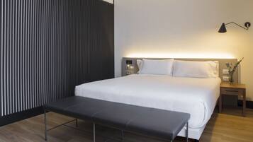 Quarto de hotel moderno com uma cama grande, um banco, uma mesa de cabeceira com abajur e uma parede com linhas verticais.