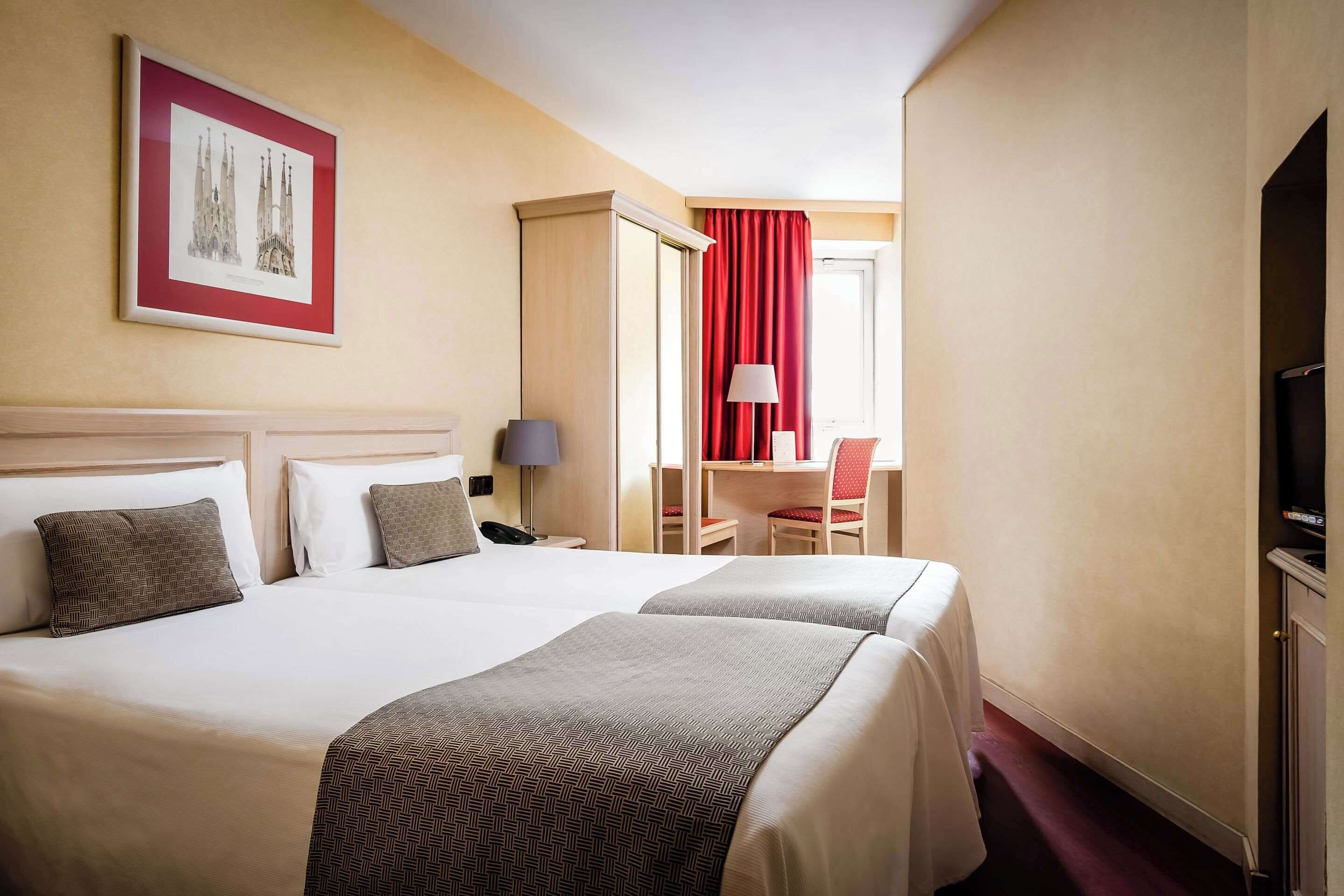 Quarto de hotel com duas camas, uma escrivaninha, uma cadeira, uma televisão e um quadro na parede.