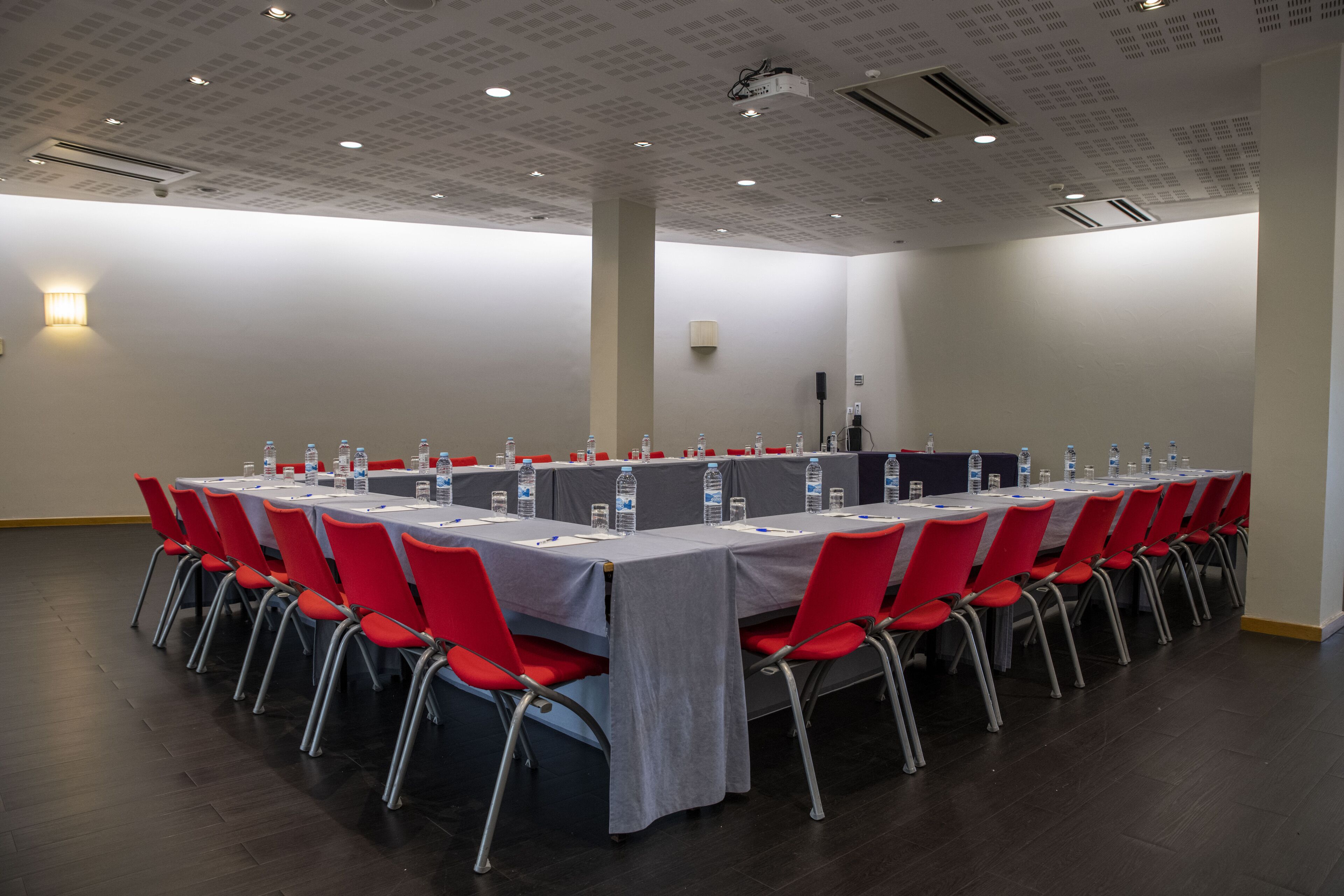 Uma sala de conferências com uma mesa comprida, cadeiras vermelhas e garrafas de água.
