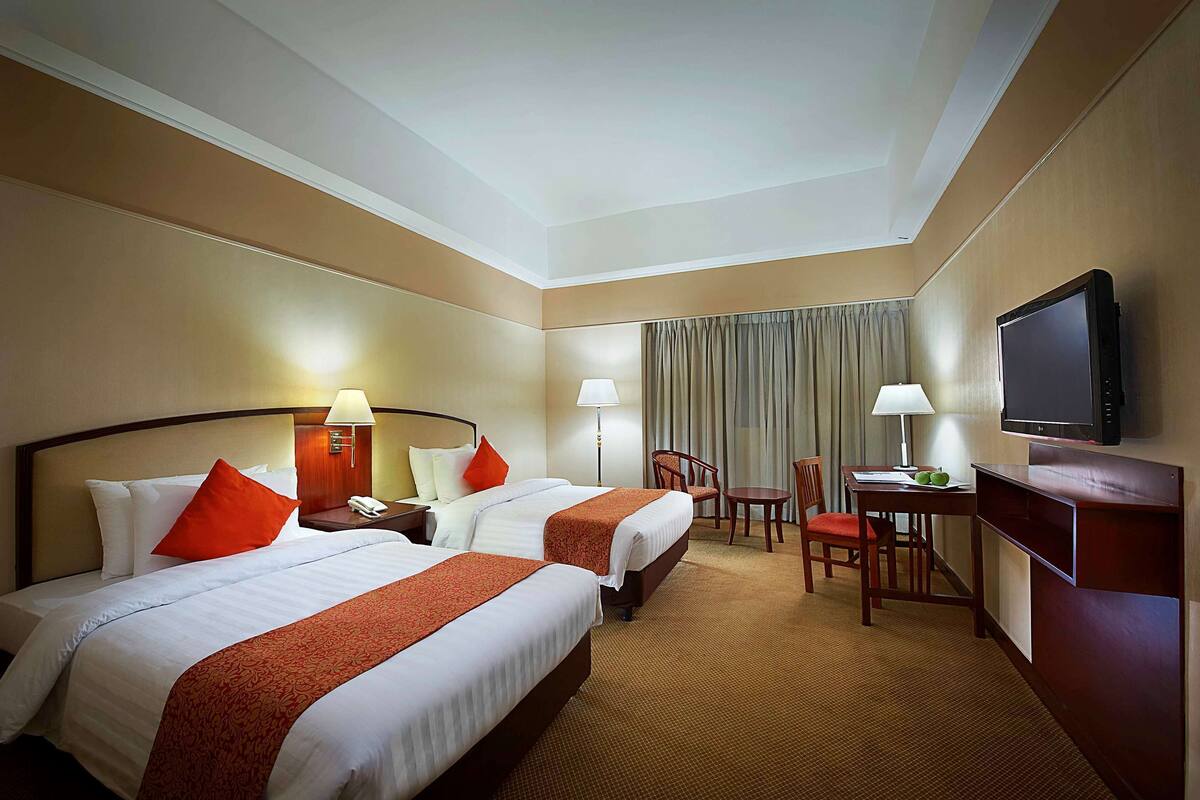 Quarto de hotel com duas camas, uma escrivaninha, uma cadeira, uma televisão e um abajur.