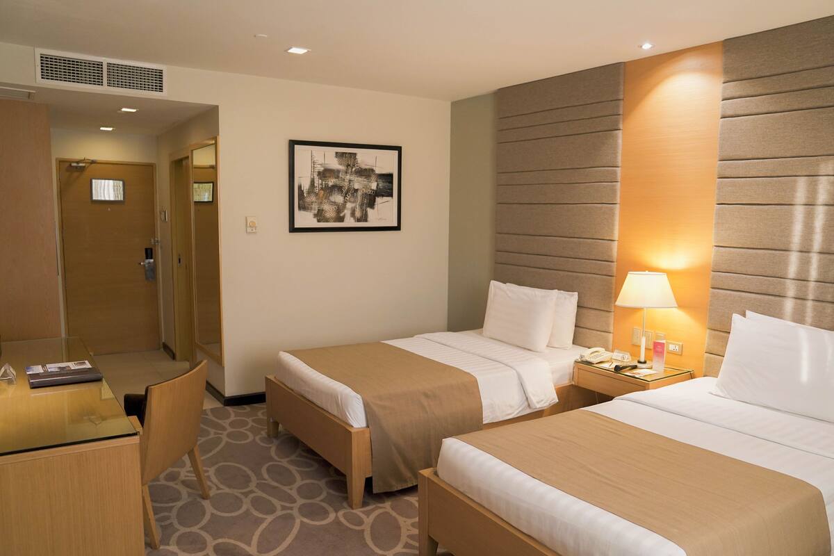 Quarto de hotel com duas camas, uma escrivaninha, uma cadeira, um quadro pendurado na parede e um abajur.