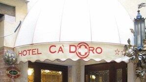Entrada de hotel com tapete vermelho, porta de vidro com emblema dourado e placa com a inscrição 'HOTEL CA'DORO'.
