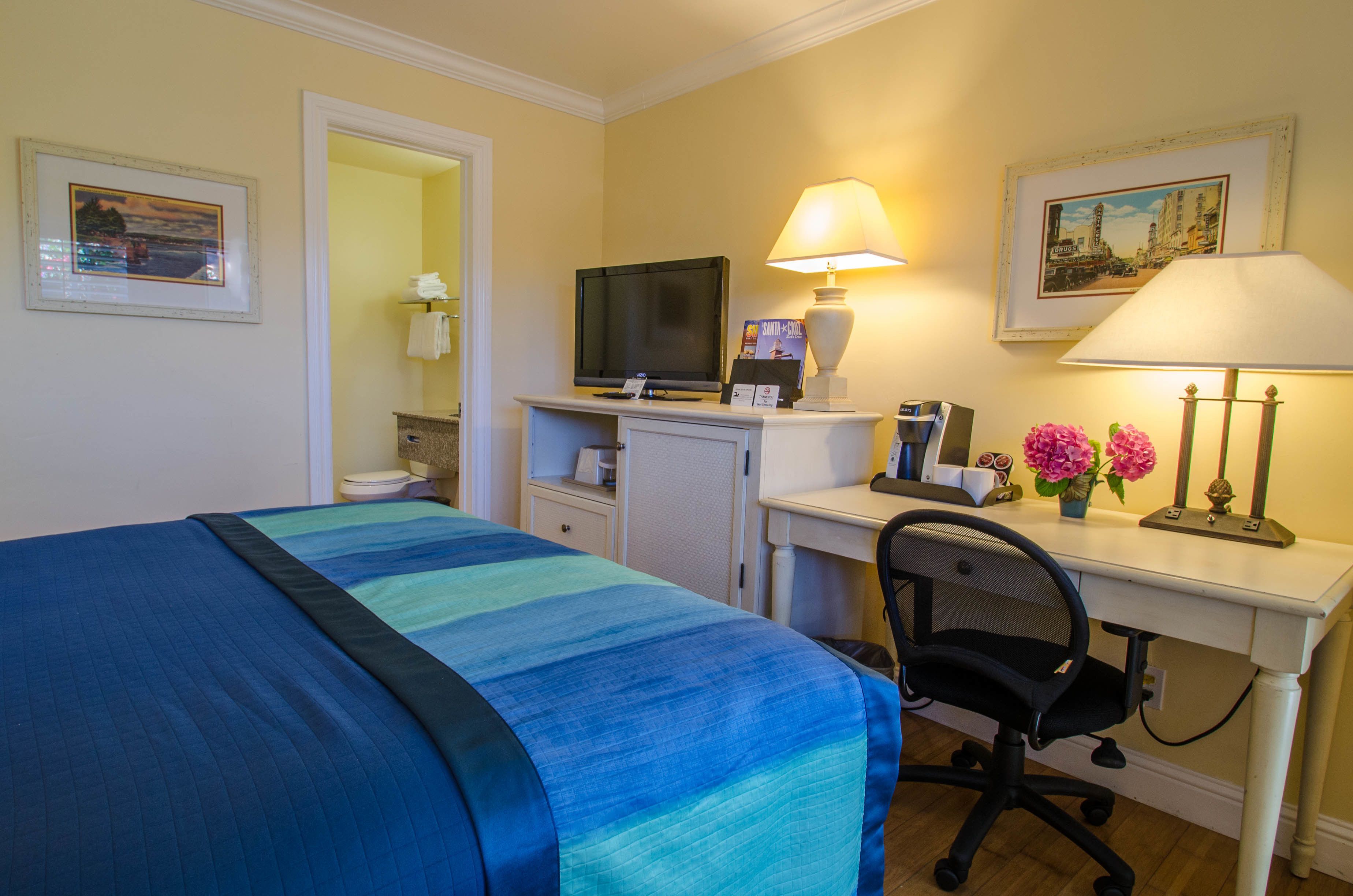 Quarto de hotel com cama, escrivaninha, cadeira, televisão, luminárias e quadros nas paredes.