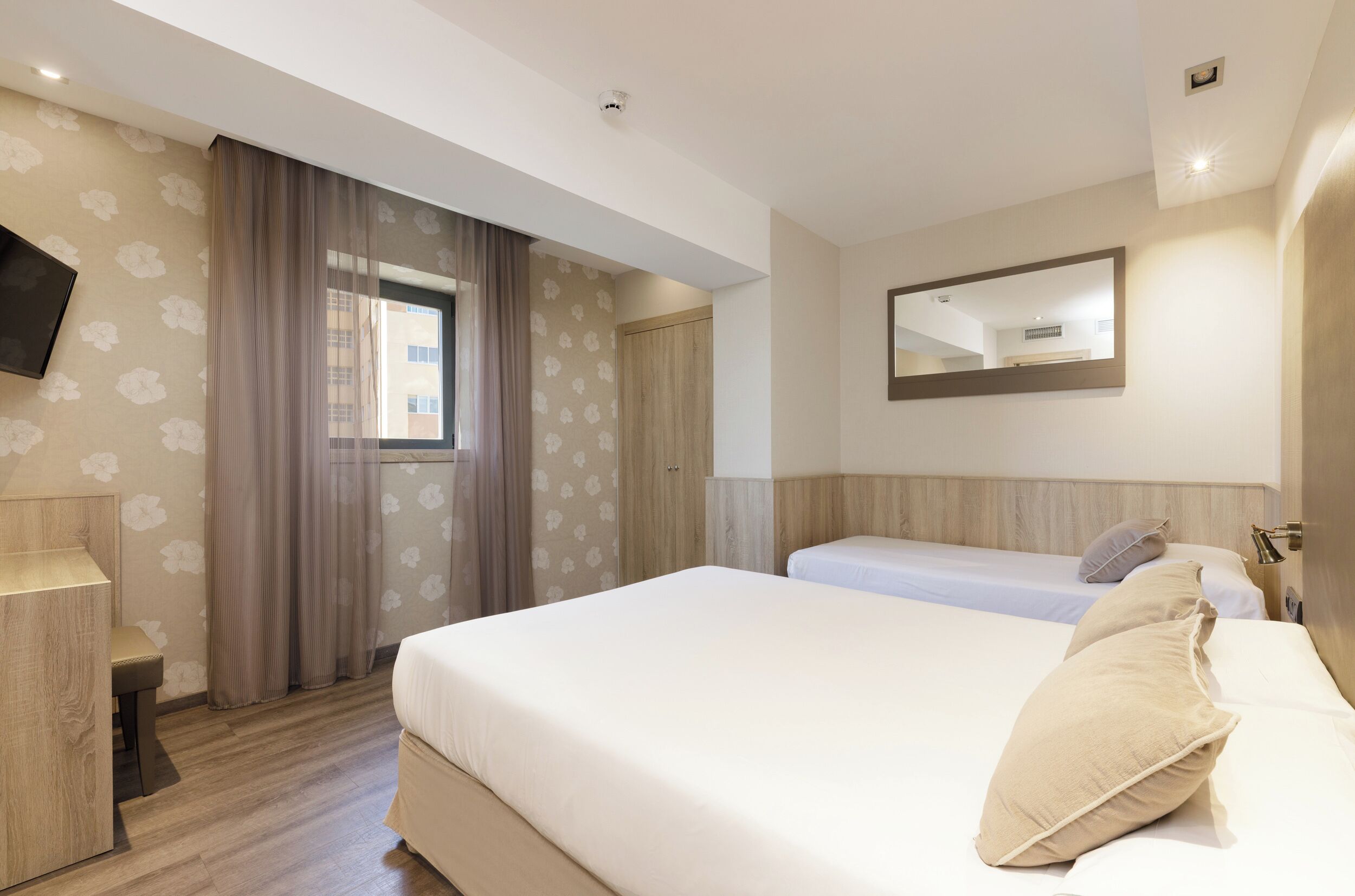 Quarto de hotel com uma cama grande, uma cama menor, uma escrivaninha e um espelho na parede.