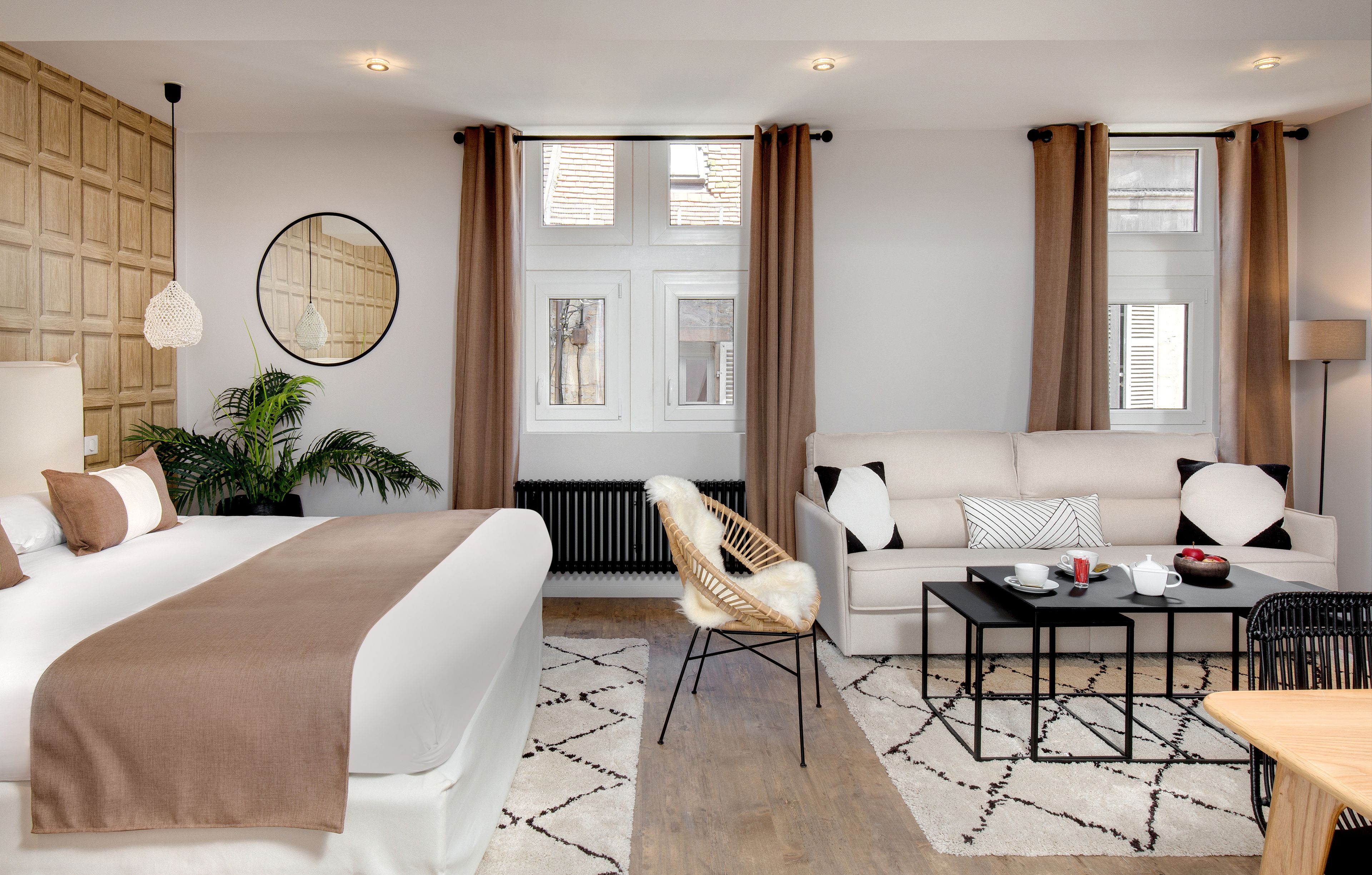 Quarto de hotel moderno com cama, sofá, mesa de jantar e uma cadeira. Há uma janela com cortinas, um radiador e uma planta em vaso.