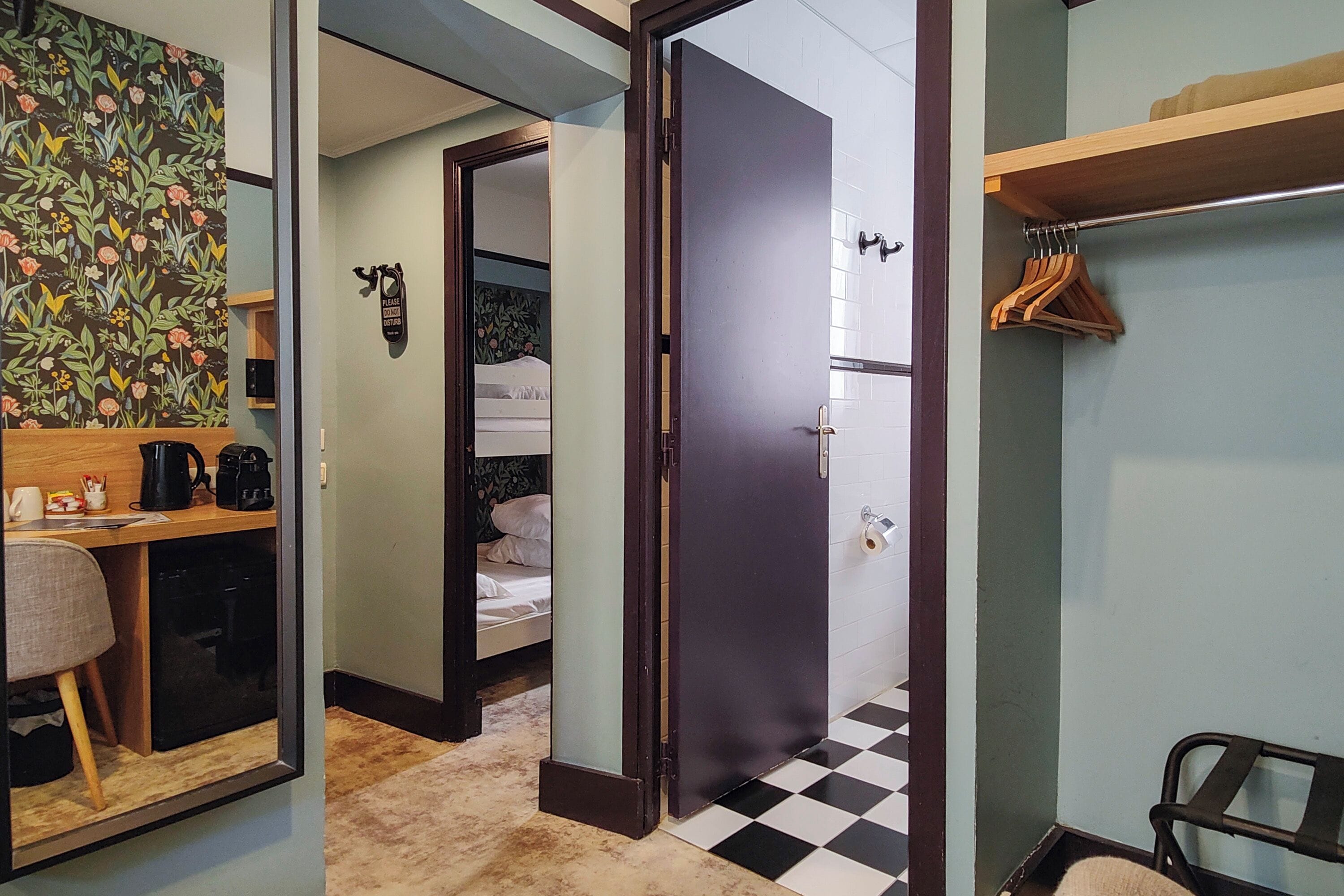Quarto de hotel com chão quadriculado, uma escrivaninha com cadeira, uma cama, guarda-roupa e um espelho.