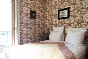 Quarto com papel de parede floral, cama com roupa de cama branca e duas molduras na parede.