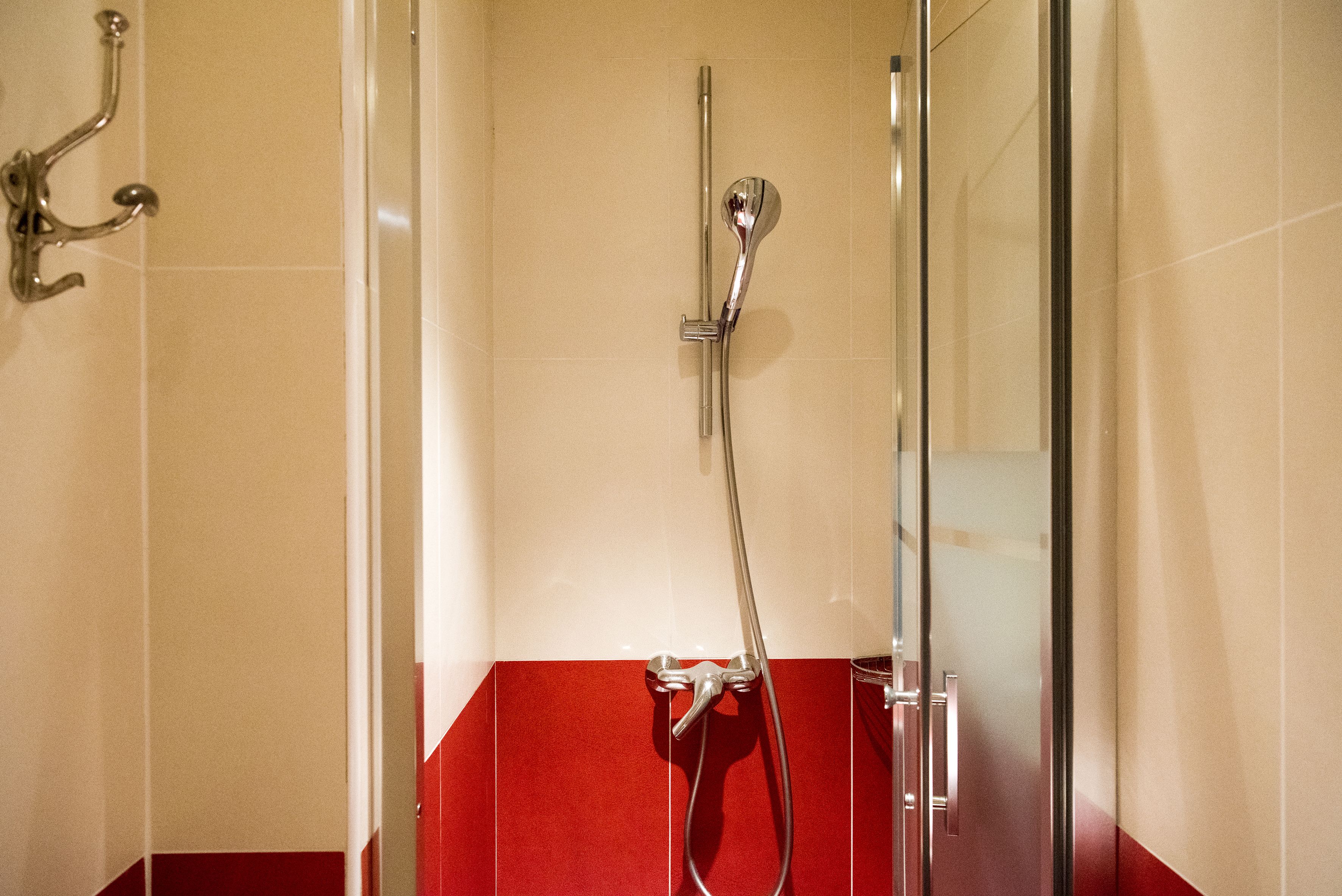 Chuveiro com chuveiro manual e torneira fixada na parede, em um espaço com paredes revestidas de azulejos vermelhos e bege.