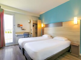 Quarto de hotel com duas camas, uma escrivaninha e uma cadeira. Há uma janela com cortinas, uma porta e uma pequena prateleira com uma chávena e um livro.
