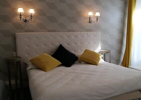 Quarto de hotel com cabeceira estofada, duas luminárias de mesa e cama com roupa de cama branca e dois travesseiros.