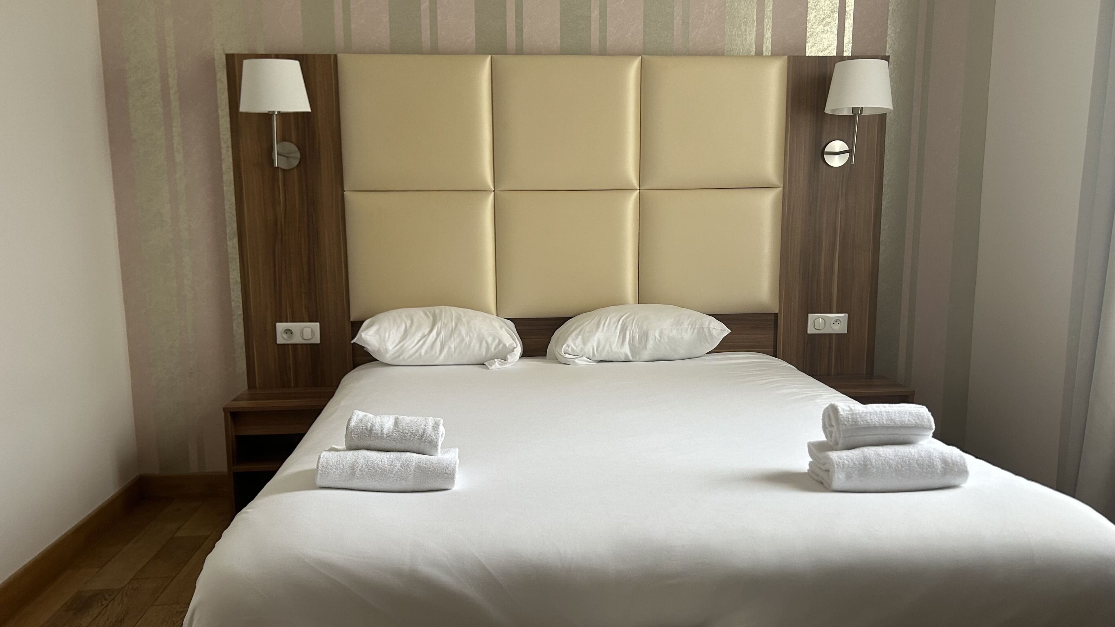 Quarto de hotel com uma cama grande, duas luminárias de cabeceira e duas toalhas dobradas na cama.