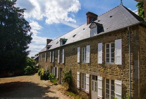 Um edifício de pedra com várias janelas e persianas, um caminho de automóveis em cascalho e vegetação ao redor.