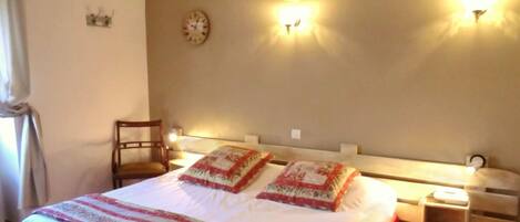 Quarto de hotel com uma cama grande, duas luminárias na parede, um relógio e uma cadeira de madeira.