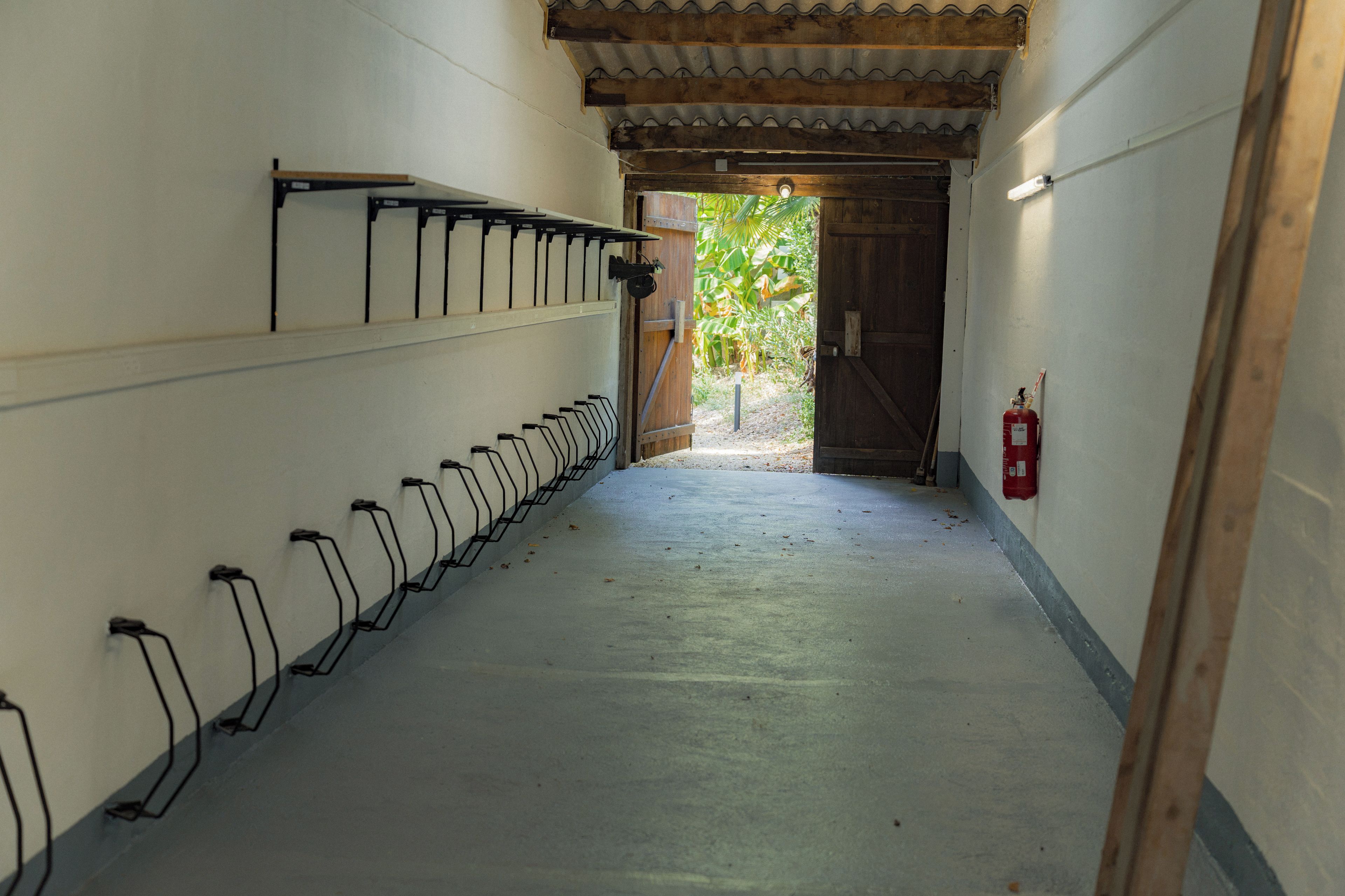 Corredor com extintor à direita, porta de madeira no final e suporte de metal com ganchos à esquerda.
