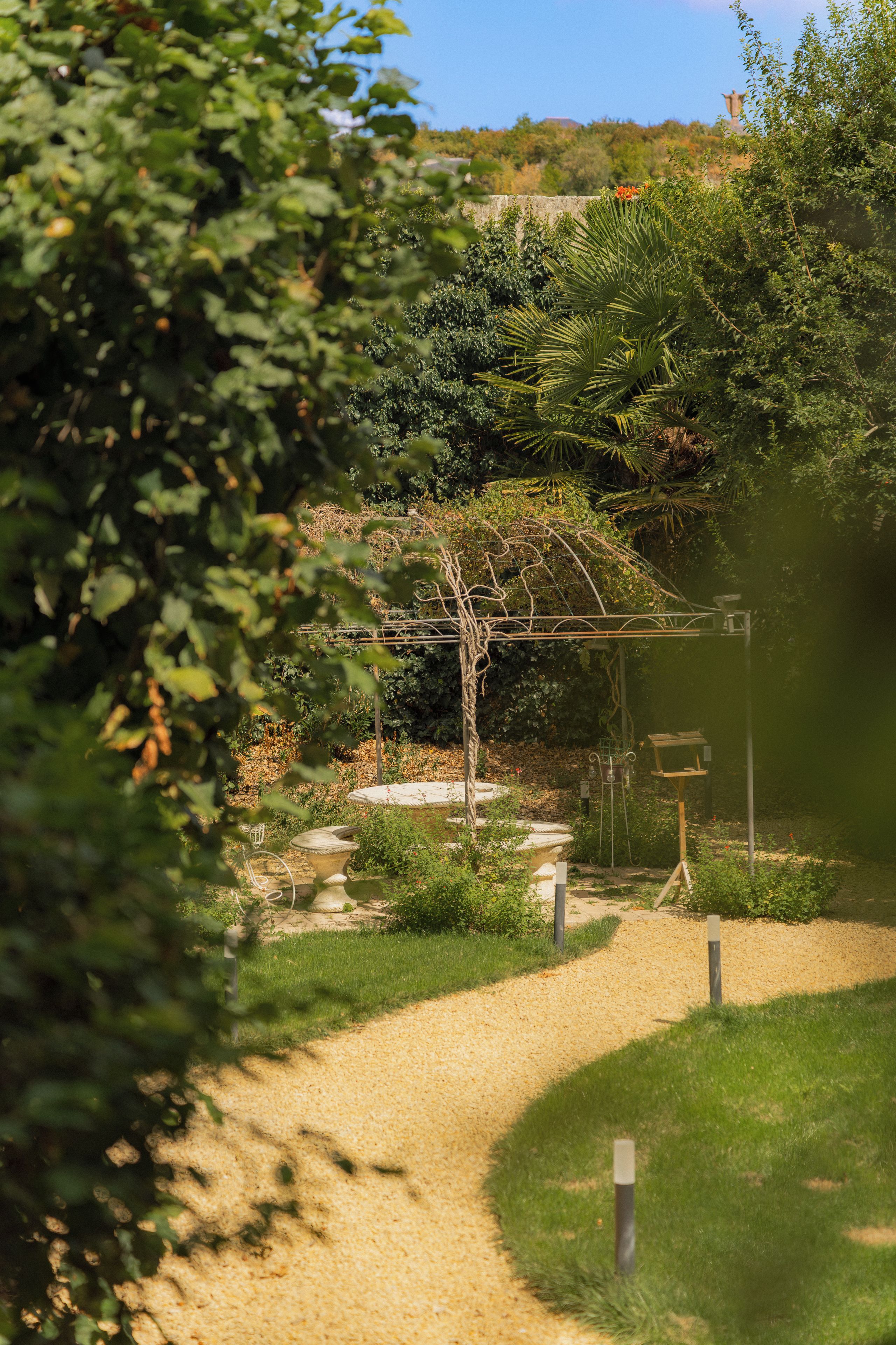 Um caminho de jardim com um banco de pedra, uma treliça de metal e um comedouro para pássaros.