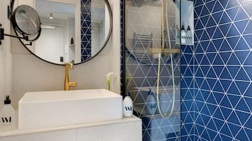 Um banheiro com pia branca, torneira dourada e espelho redondo. Há um chuveiro com divisória de vidro, chuveiro manual e um porta-toalhas fixado na parede.