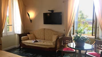 Quarto com sofá, televisão na parede, mesa pequena com cadeiras e vista para o exterior pela janela.