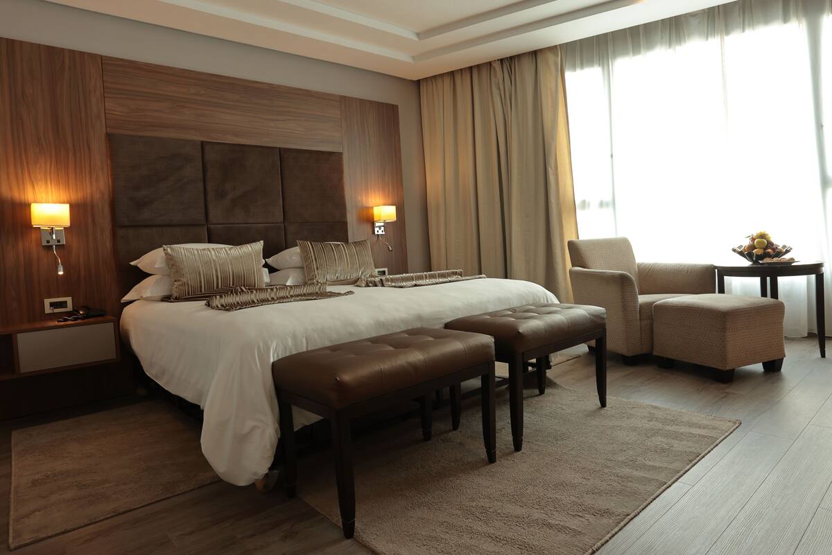 Quarto de hotel com uma cama grande, duas luminárias de cabeceira, um painel de cabeceira de madeira, um banco marrom, uma cadeira e uma mesinha com frutas.