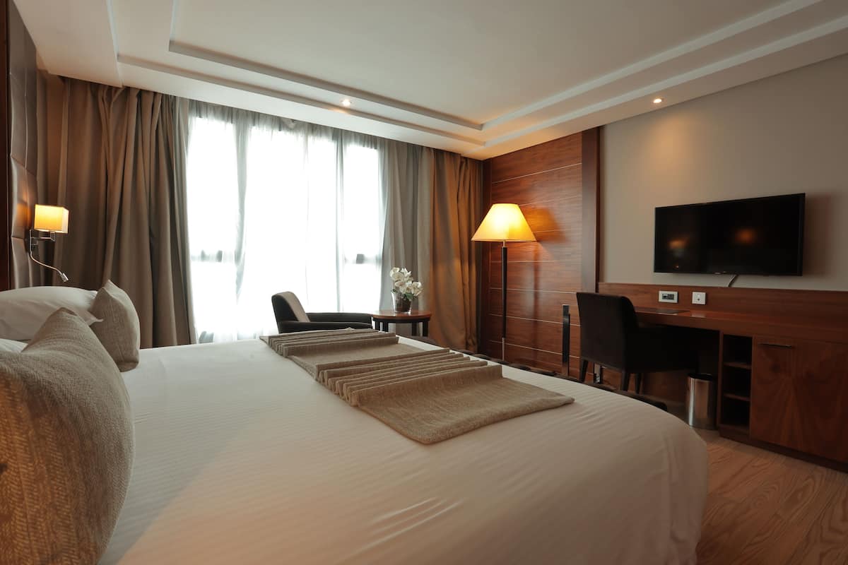 Quarto de hotel com uma cama grande, uma escrivaninha, uma cadeira, uma televisão e uma janela com cortinas.