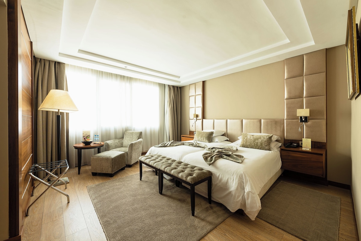 Quarto de hotel com uma cama grande, um banco, uma escrivaninha e uma cadeira.