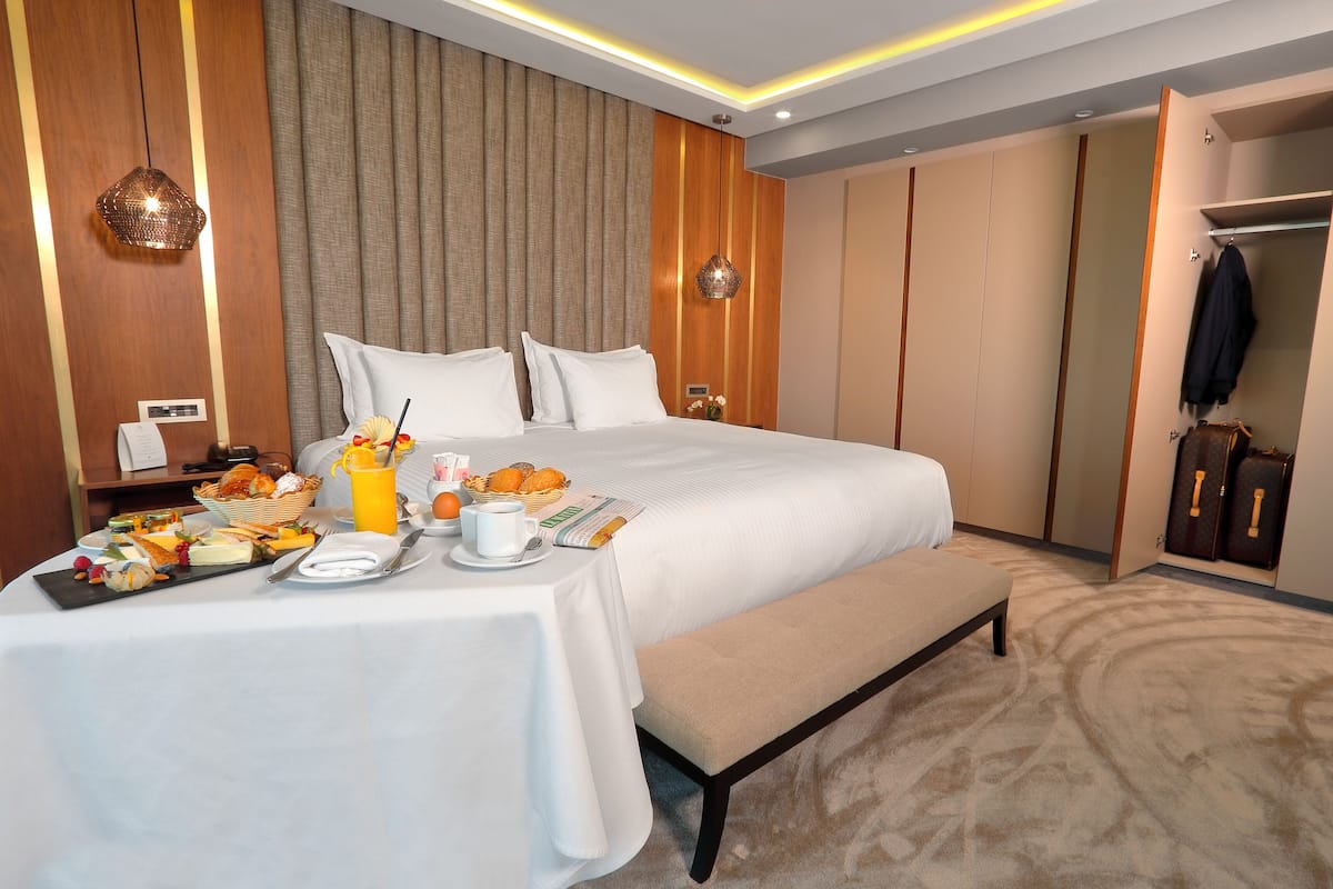 Quarto de hotel com cama bem arrumada, bandeja de café da manhã com doces e frutas, mesa de cabeceira com abajur e guarda-roupa com malas.