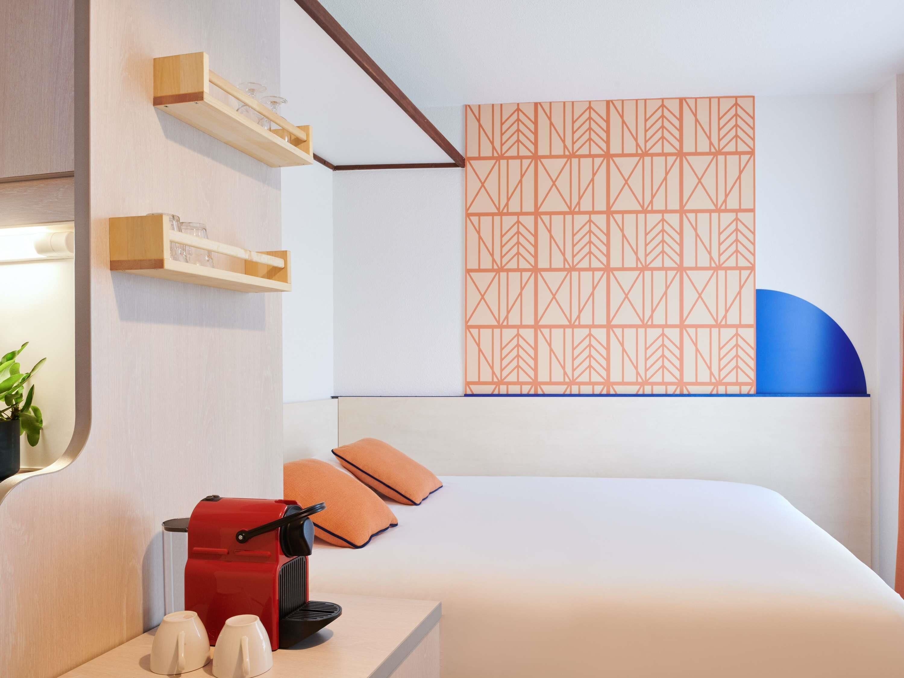 Quarto de hotel moderno com cama, cafeteira, pequena mesa com copos e um painel decorativo na parede.