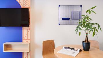 Quarto moderno com uma mesa redonda de madeira, uma planta em vaso, uma televisão de tela plana na parede e uma estante azul com padrão geométrico.