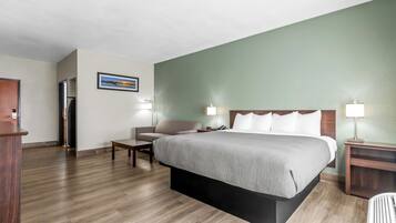 Quarto de hotel com cama, uma cadeira, escrivaninha, abajur e um quadro na parede.