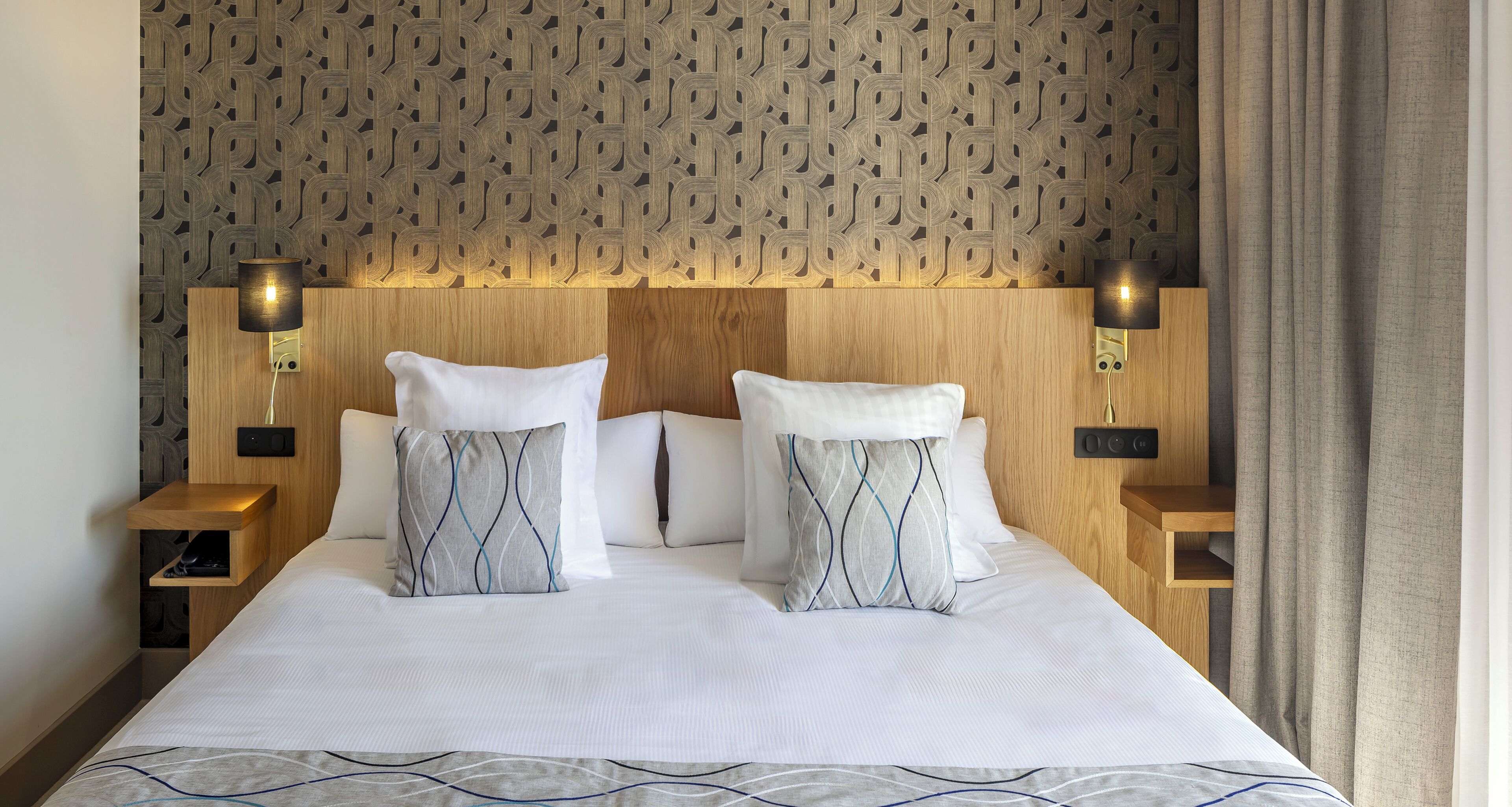 Cama bem arrumada com lençóis brancos e almofadas decorativas, cabeceira de madeira e abajures laterais.