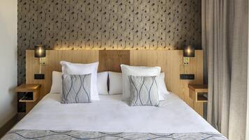 Uma cama bem arrumada com lençóis brancos e duas almofadas decorativas, com duas mesas de cabeceira e abajures de cada lado, em um quarto com papel de parede estampado e cabeceira de madeira.