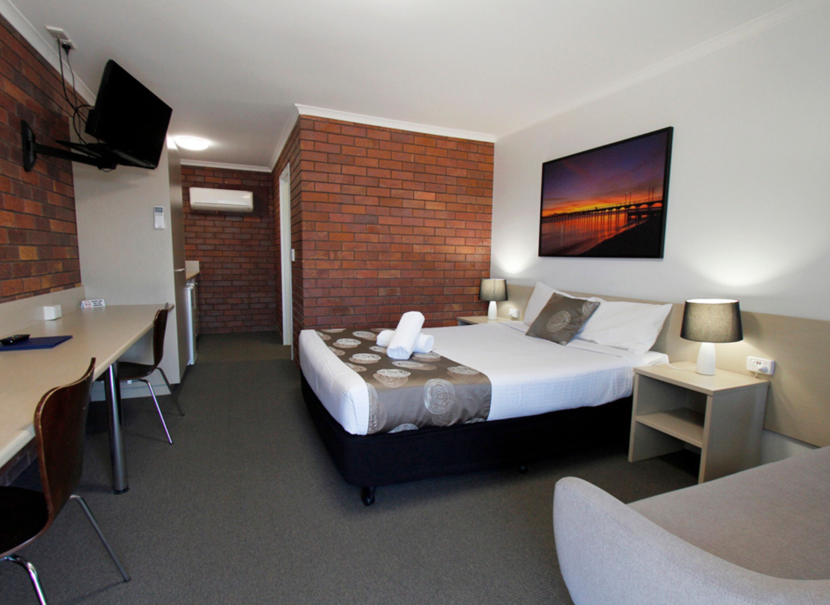 Quarto de hotel com cama, escrivaninha, cadeira, televisão e um quadro na parede.
