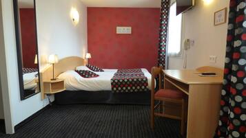 Quarto de hotel com cama, luminárias laterais, escrivaninha com cadeira, telefone e espelho.