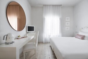 Quarto de hotel com um espelho redondo, uma escrivaninha com computador, uma cama e uma janela com cortinas leves.