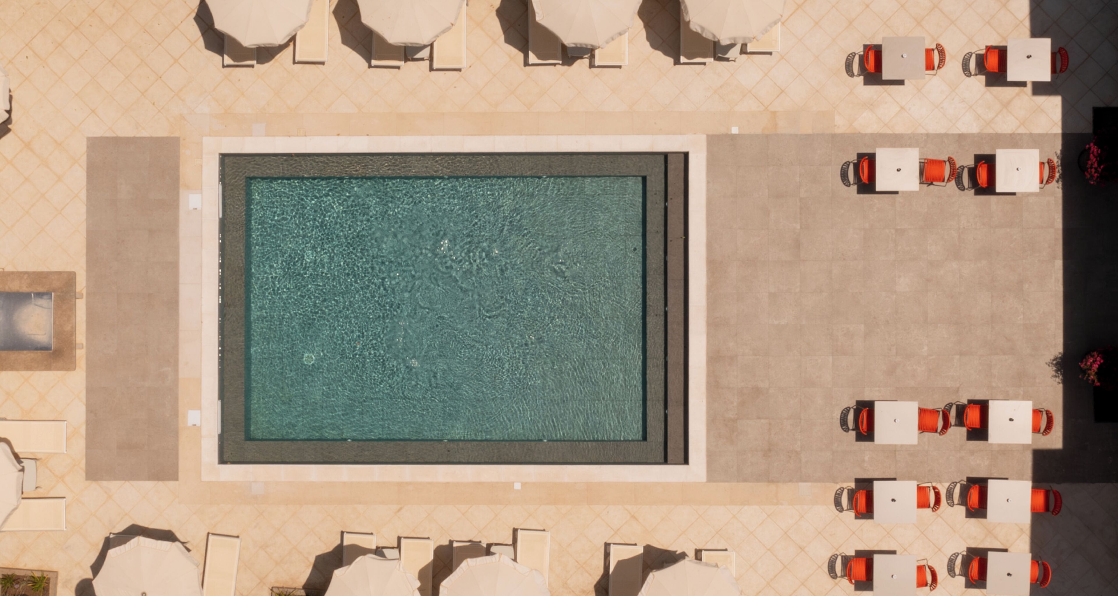 Vista aérea de uma área de piscina com um deck ladrilhado, espreguiçadeiras e mesas.