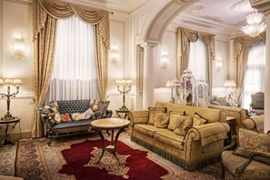 Uma sala de estar luxuosa com um sofá azul, um sofá bege e um tapete vermelho. Há duas luminárias em mesas laterais e um candelabro.