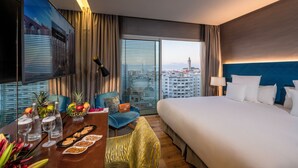 Quarto de hotel com uma cama grande, uma mesa de jantar com frutas e copos, uma TV e vista para a cidade.