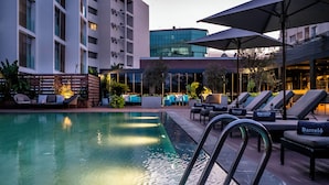 Área da piscina com espreguiçadeiras, piscina e um edifício ao fundo.