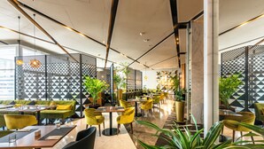 Restaurante moderno com tetos altos, paredes com padrão geométrico e diversas opções de assento, incluindo cadeiras amarelas e sofás verdes.