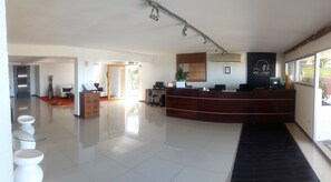 Uma área de recepção de hotel com um balcão de madeira, um computador e uma planta. Há um aviso na parede e uma janela com vista para o exterior.