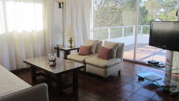 Quarto de hotel com sofá, uma mesa pequena, luminária, televisão e varanda com vista para árvores.