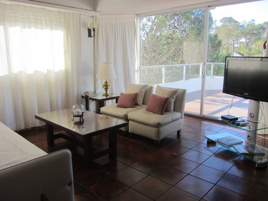 Quarto de hotel com sofá, uma mesa pequena, luminária, televisão e varanda com vista para árvores.