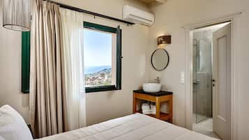 Quarto com cama, janela com cortinas, mesa de cabeceira e pia, além de vista para a cidade.