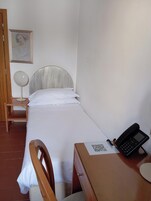 Uma cama de solteiro com lençóis brancos, uma mesa de cabeceira com luminária, uma escrivaninha com telefone e um quadro na parede.